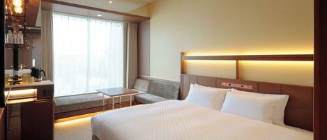 Quarto de hotel com uma cama grande, área de estar, escrivaninha com cafeteira e janela com cortinas.