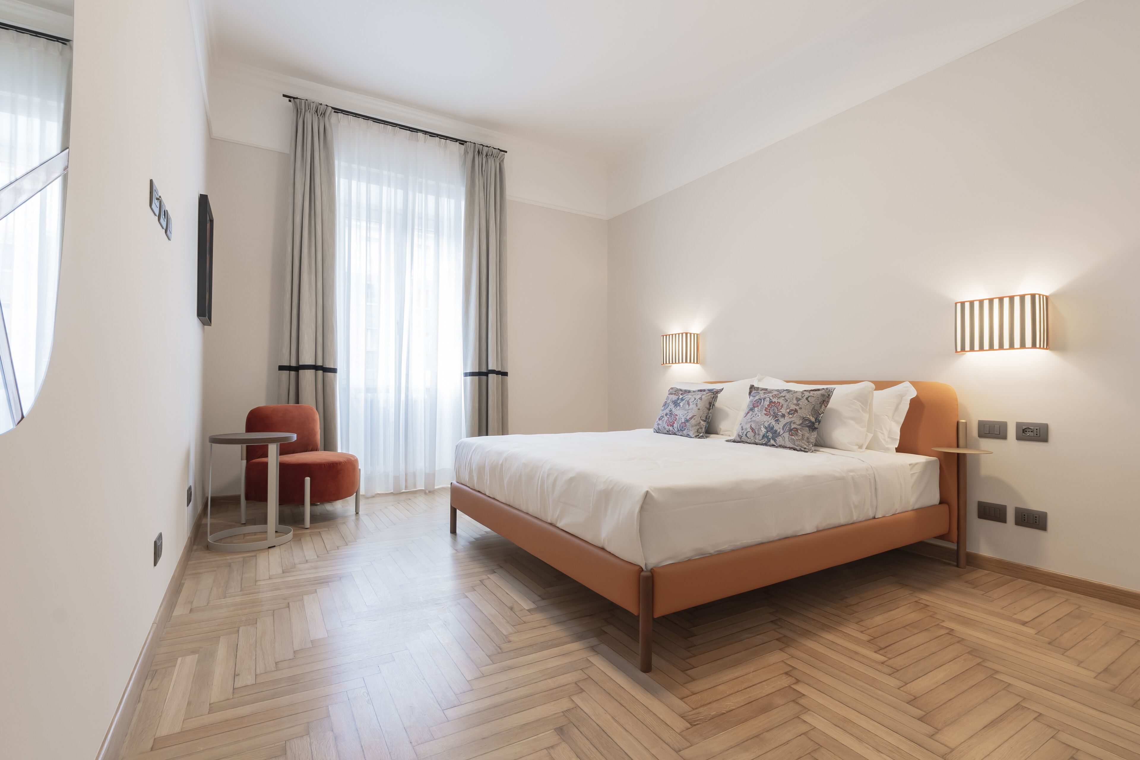 Quarto de hotel moderno com uma cama grande, uma escrivaninha e uma cadeira. Há duas luminárias fixadas na parede e uma janela com cortinas.