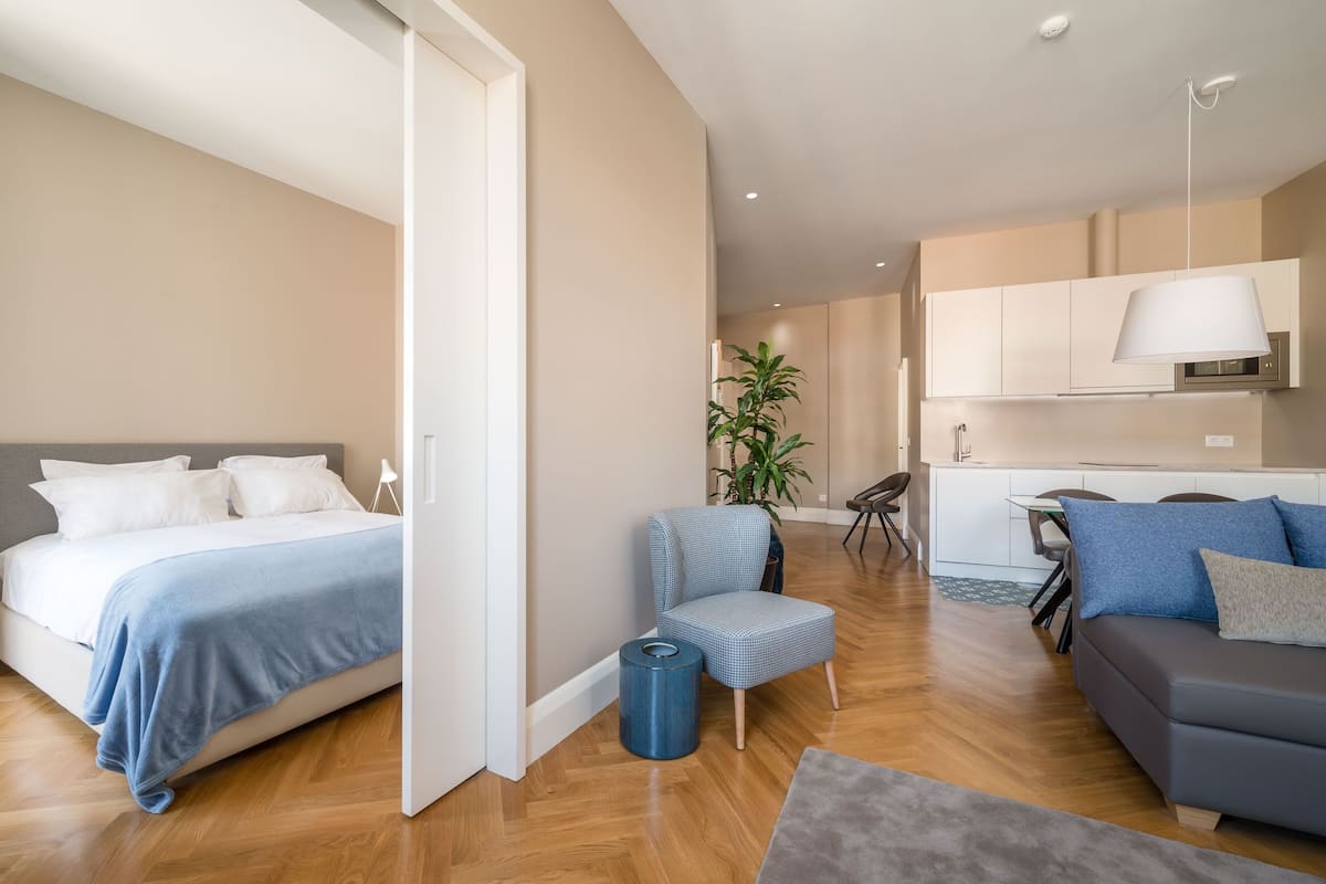Quarto de hotel moderno com cama, área de estar com sofá e poltrona, mesa pequena e planta em vaso.
