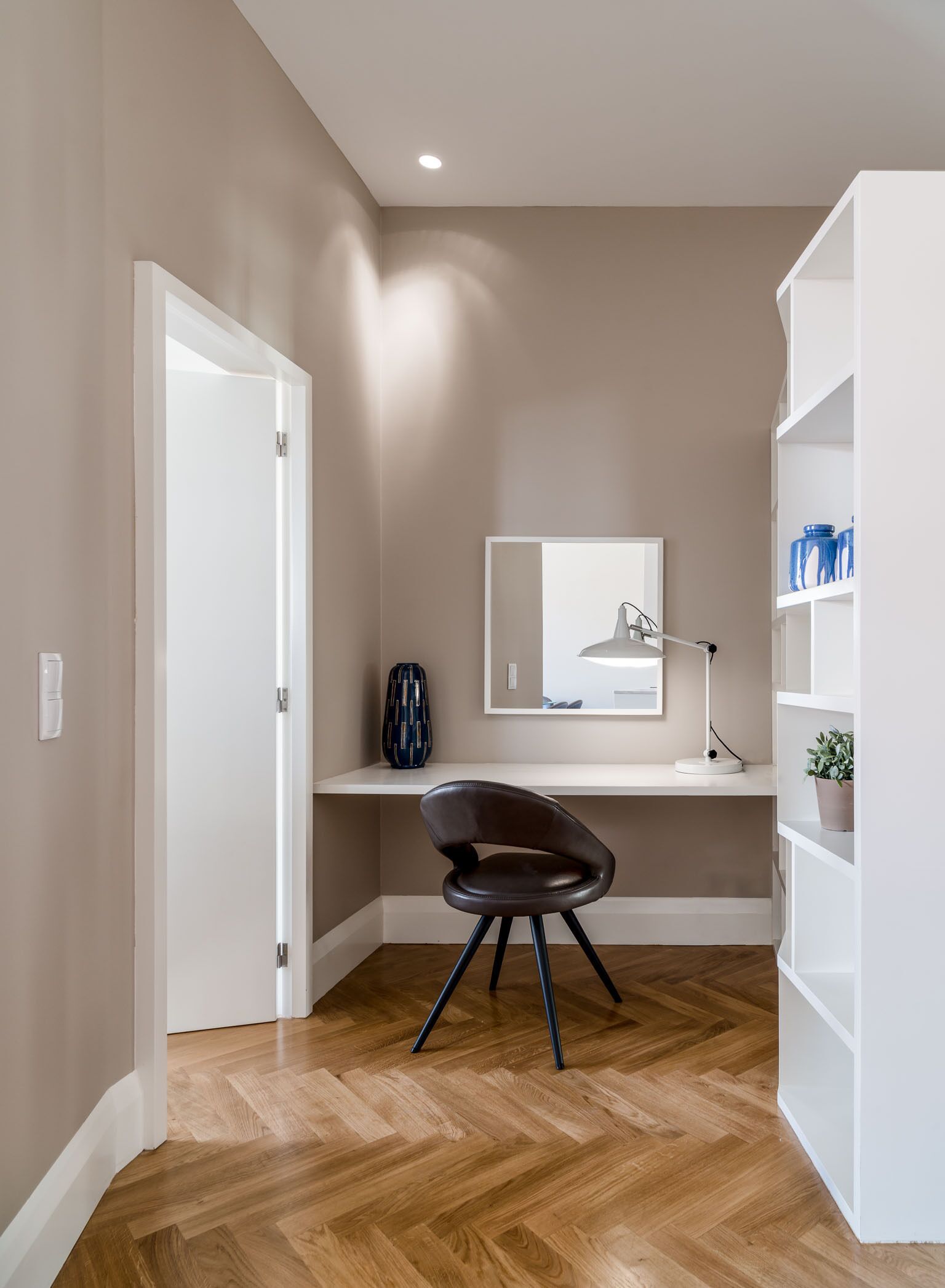 Quarto moderno e bem iluminado com piso de madeira em padrão herringbone, uma escrivaninha com espelho, uma cadeira e uma estante branca com objetos decorativos.