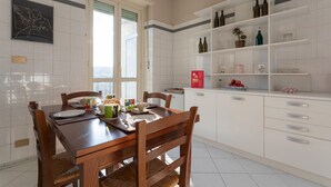Uma área de refeições com mesa e cadeiras de madeira, uma cozinha com armários e prateleiras brancas, e uma janela com vista.