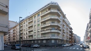 Um edifício residencial com vários andares, varandas, faixa de pedestres e carros estacionados.