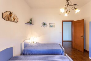 Quarto com duas camas, uma mesa de cabeceira, luminária na parede, uma porta e um corredor que dá para outro cômodo.