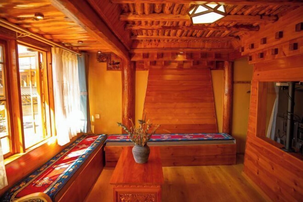 Interior de madeira com um banco, uma mesa com um vaso e uma janela com cortinas.