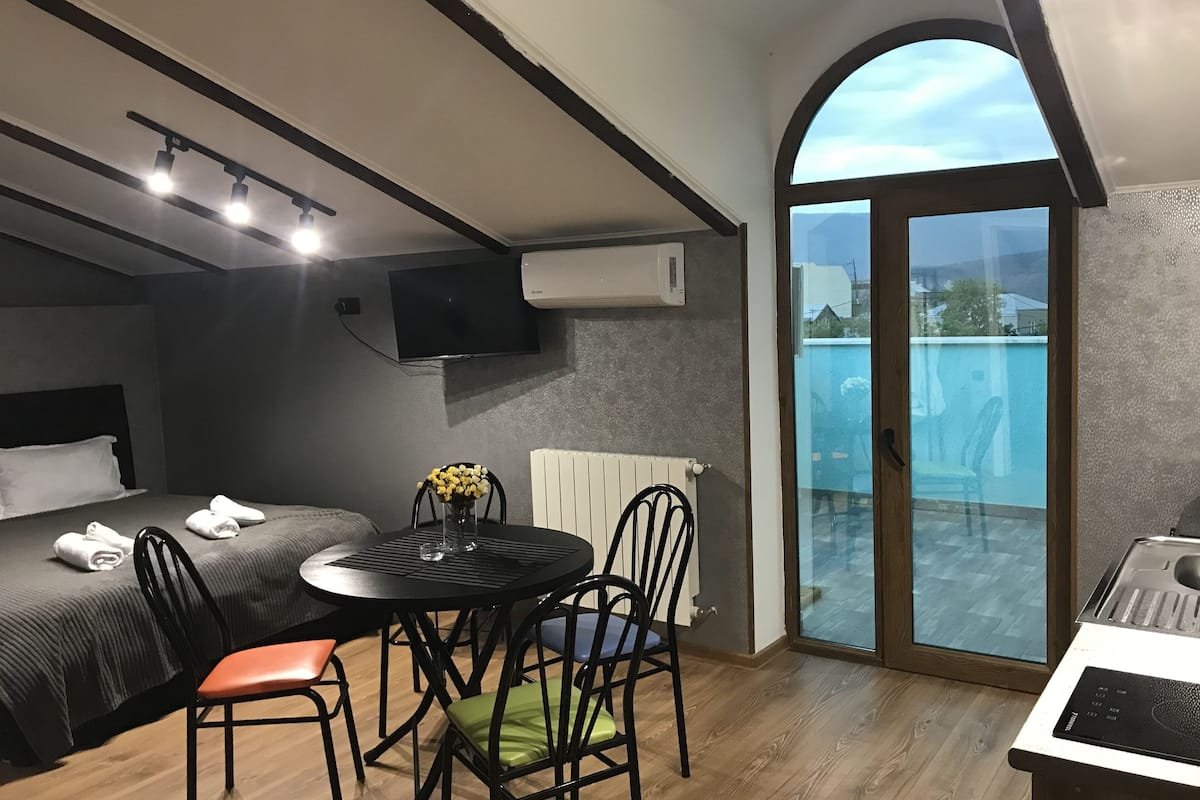 Quarto de hotel compacto com cama, mesa de jantar com cadeiras, pequena cozinha e televisão na parede. Porta de vidro desliza para uma varanda.