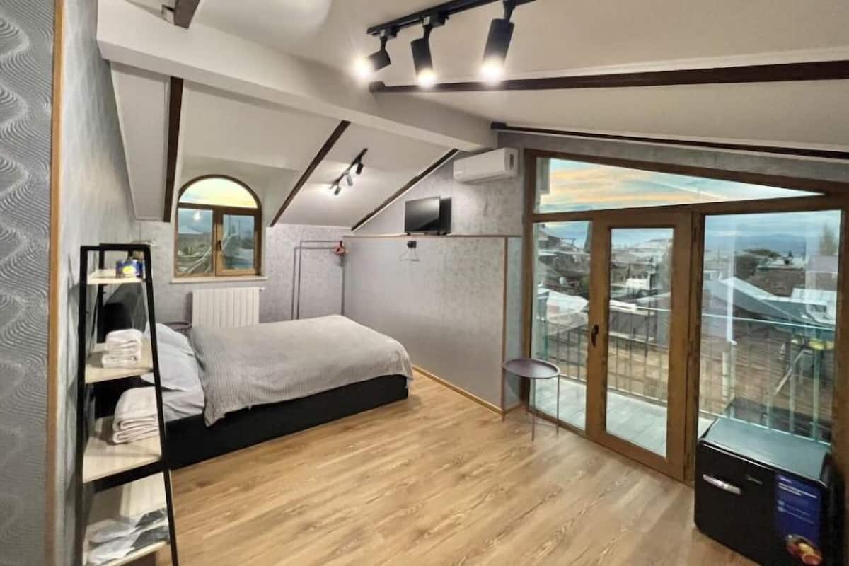 Quarto com cama, prateleira com toalhas dobradas, mesa pequena e grande janela com vista para edifícios.