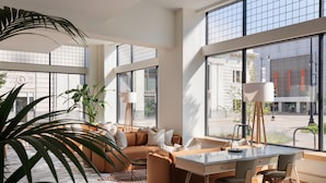 Uma sala de estar moderna com um sofá, uma mesa de vidro e cadeiras. Há janelas amplas e vista para a rua.