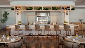 Uma cozinha moderna com ilha central, banquetas de bar e área de estar com mesas redondas.