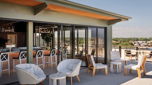 Um bar no terraço com área externa, incluindo cadeiras e mesas de vime, e vista para a paisagem urbana.