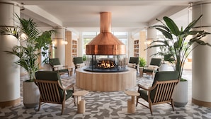 Um lobby moderno com lareira central, áreas de estar e grandes vasos de plantas.