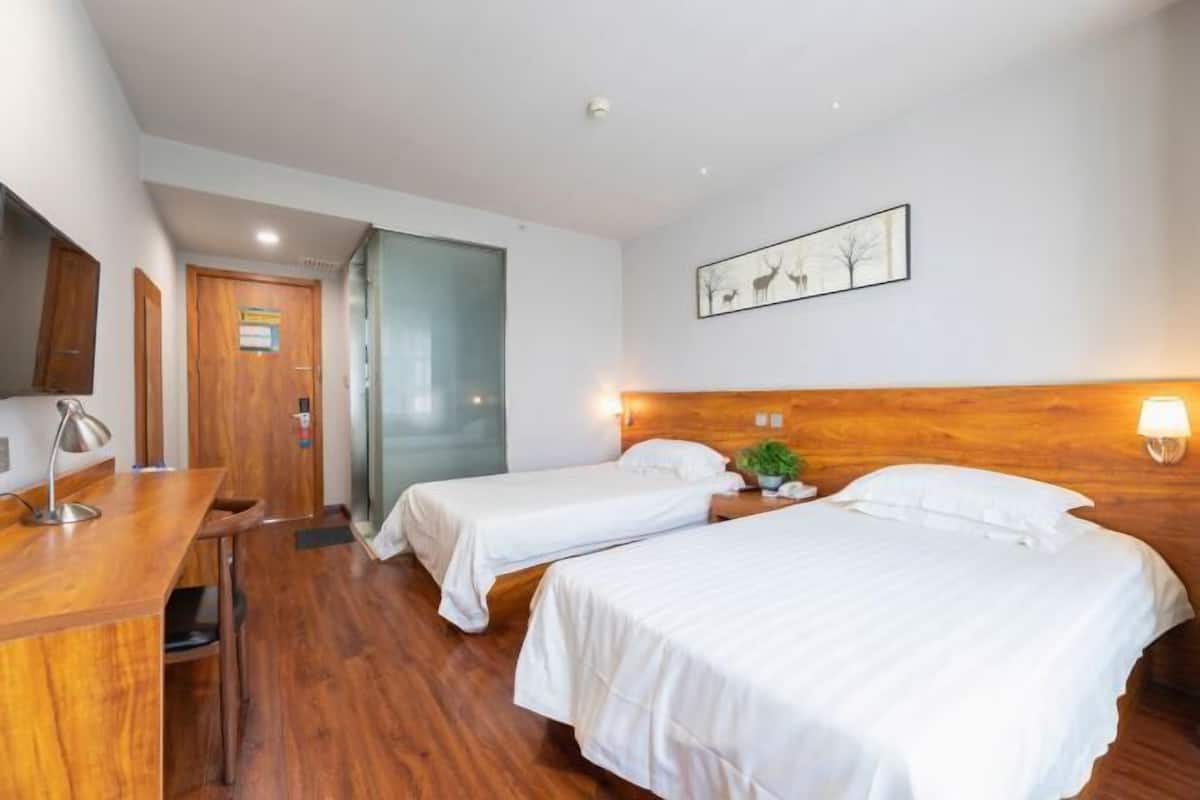 Quarto de hotel com duas camas de solteiro, uma escrivaninha de madeira com uma luminária, uma mesinha e uma televisão fixada na parede.