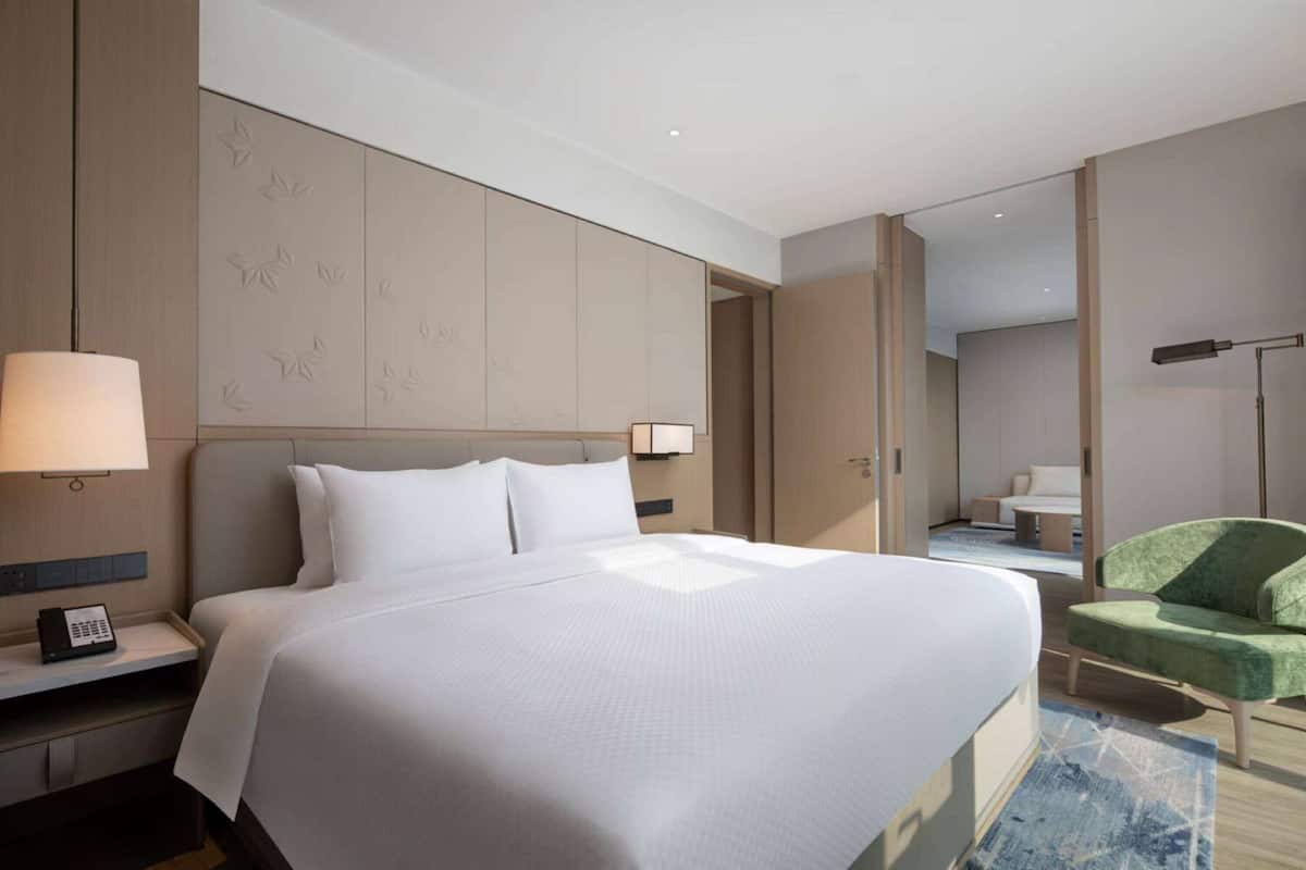 Quarto de hotel moderno com uma cama grande, uma poltrona verde, uma mesa de cabeceira com abajur e um espelho.