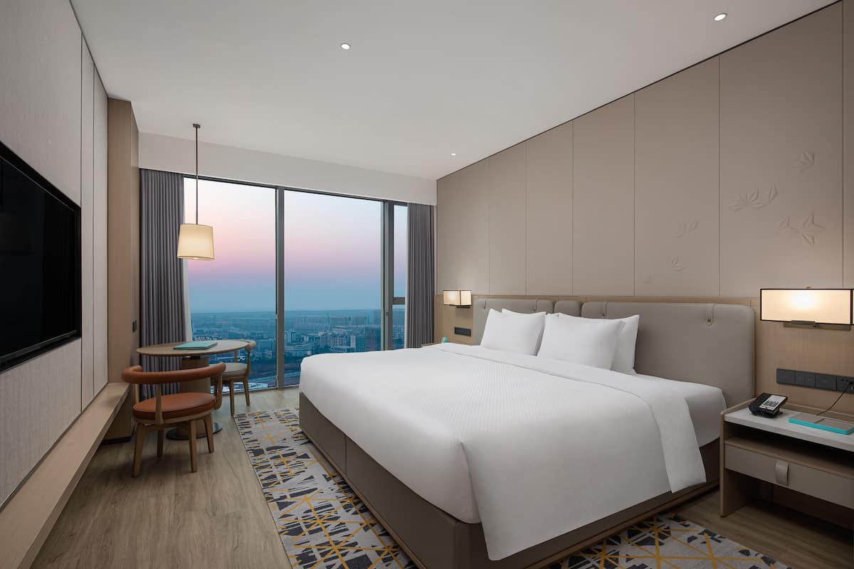 Quarto de hotel moderno com uma cama grande, uma escrivaninha com luminária, uma cadeira e uma televisão.