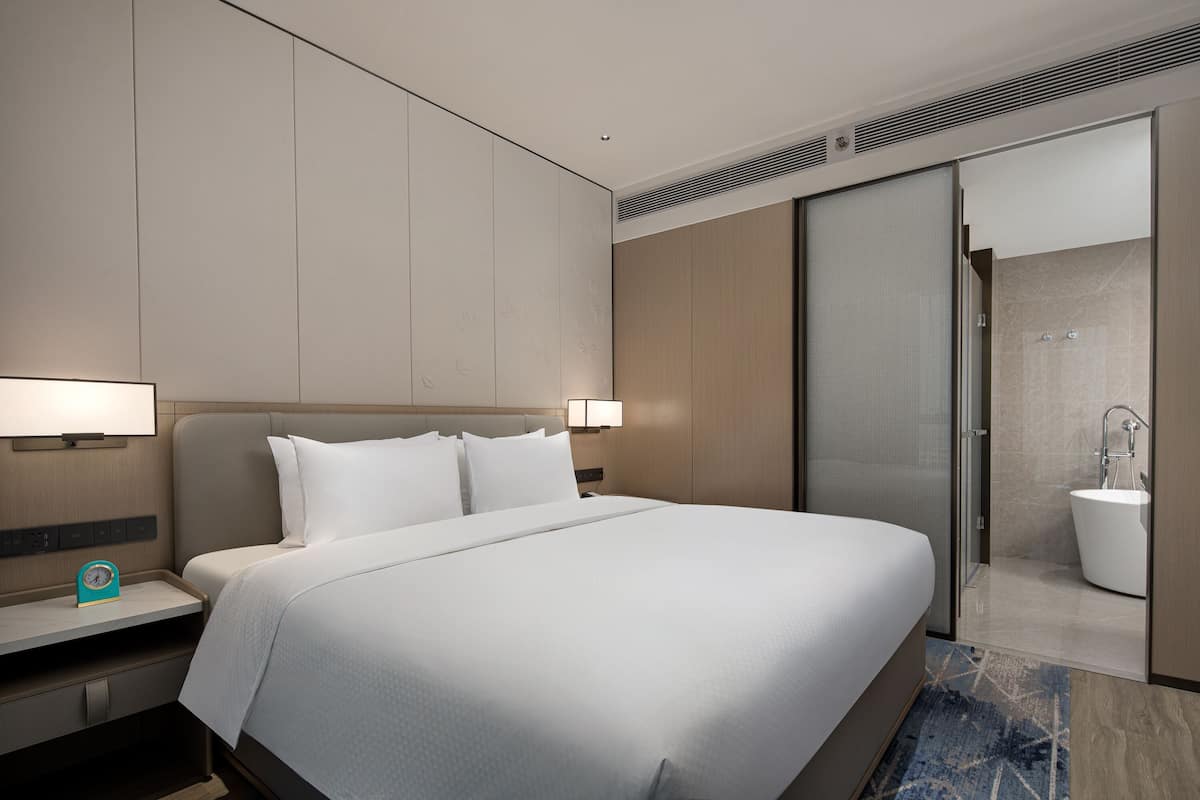 Quarto de hotel moderno com uma cama grande, mesas de cabeceira com abajures, banheiro com banheira e um armário com portas de correr.