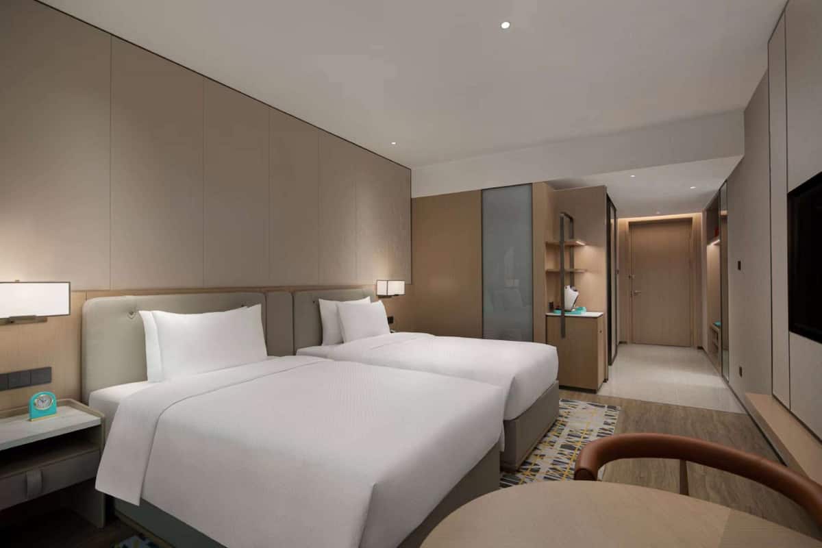 Quarto de hotel com duas camas, uma escrivaninha, uma cadeira, televisão e banheiro visível por uma porta.