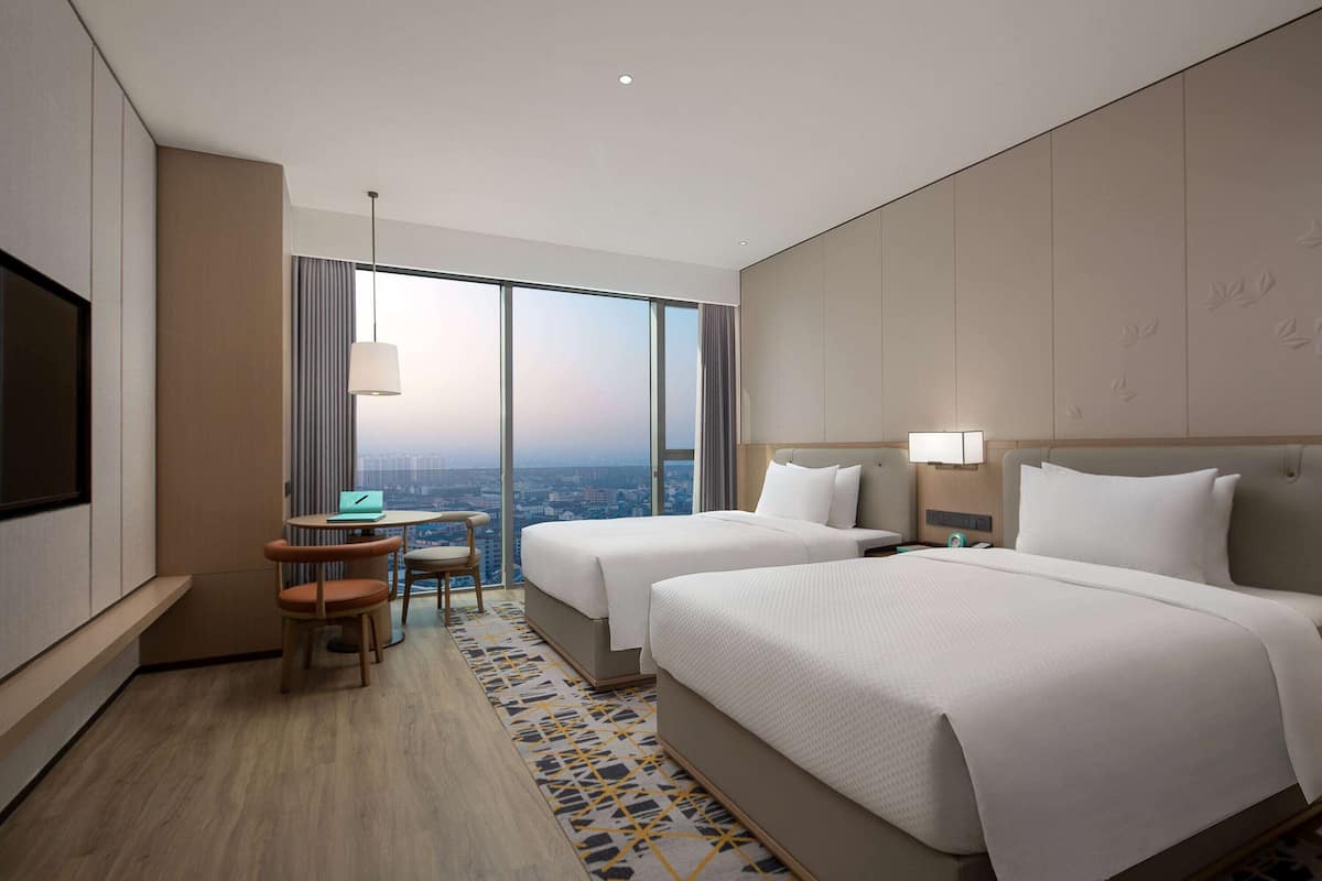 Quarto de hotel com duas camas, uma escrivaninha com um laptop, uma cadeira e uma janela grande com vista para a cidade.