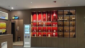 Uma área de recepção com um quiosque digital, uma vitrine vermelha com diversos itens e um painel de parede em madeira.