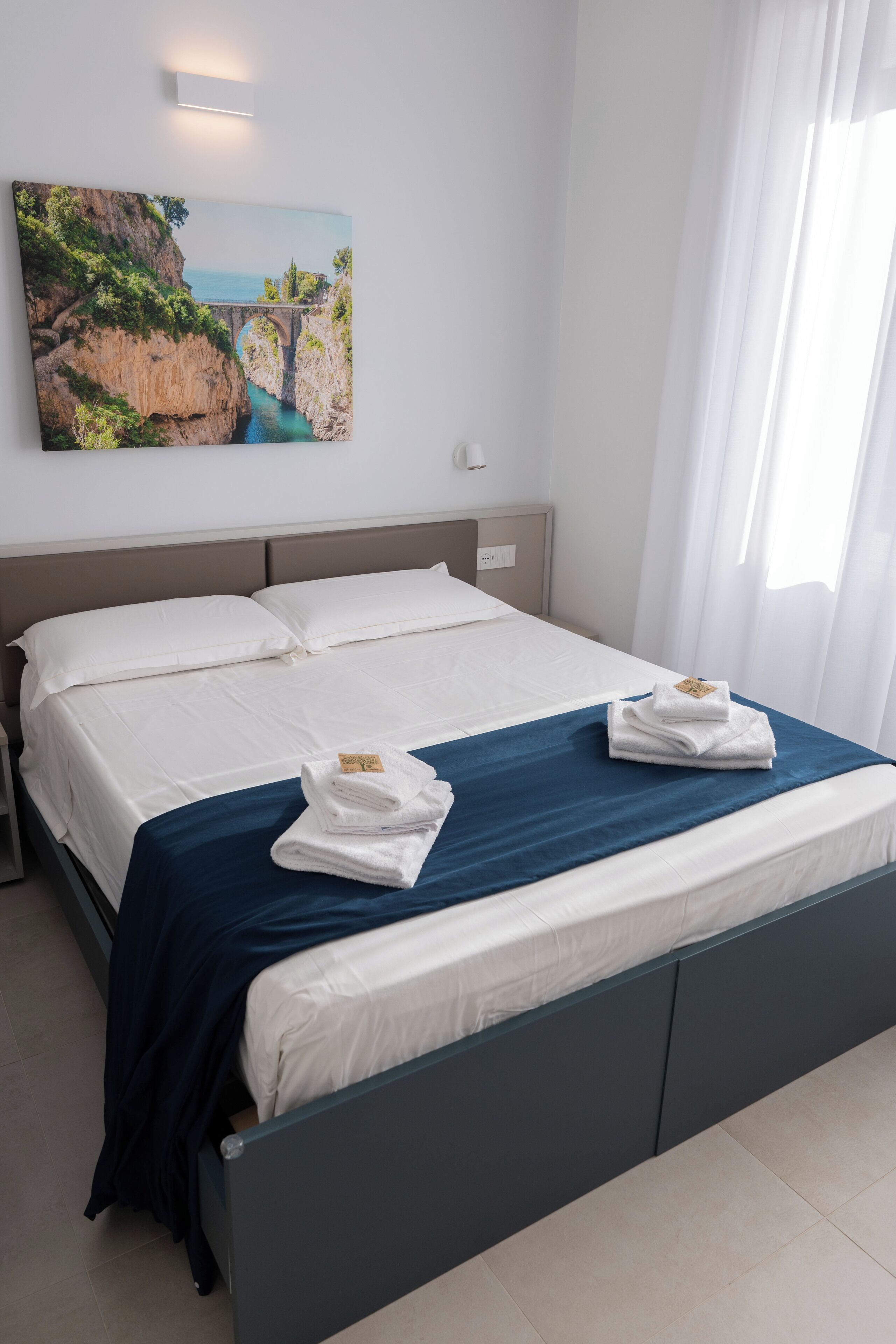 Uma cama bem arrumada com lençóis brancos e colcha azul, um quadro com uma paisagem costeira na parede e uma janela com cortinas finas.