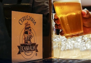 Mão segurando um copo de cerveja ao lado de uma garrafa com a etiqueta 'Cerveja KABRALA Porto Seguro Brasil'.