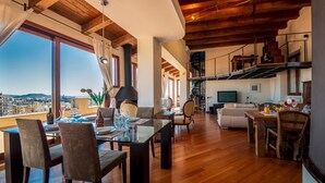 Uma sala de estar espaçosa com uma mesa de jantar, cadeiras e um sofá. Há uma televisão e uma lareira. A sala possui grandes janelas com vista para a cidade.