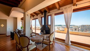 Um quarto amplo com teto alto, vigas de madeira, lareira, área de refeições com mesa e cadeiras, e janelas grandes com vista para a cidade.