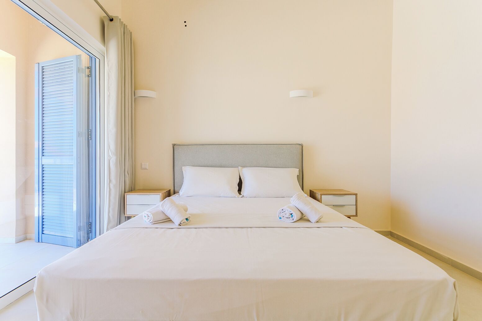 Quarto de hotel com uma cama grande, duas mesas de cabeceira, uma janela com persianas e um ponto de luz fixado na parede.