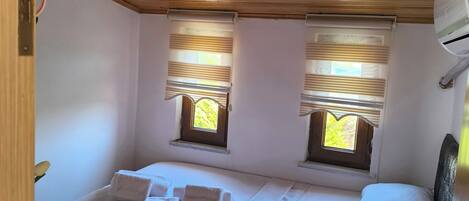 Uma cama de casal com roupa de cama branca, duas janelas com persianas, um ventilador de teto e um ar condicionado fixado na parede.
