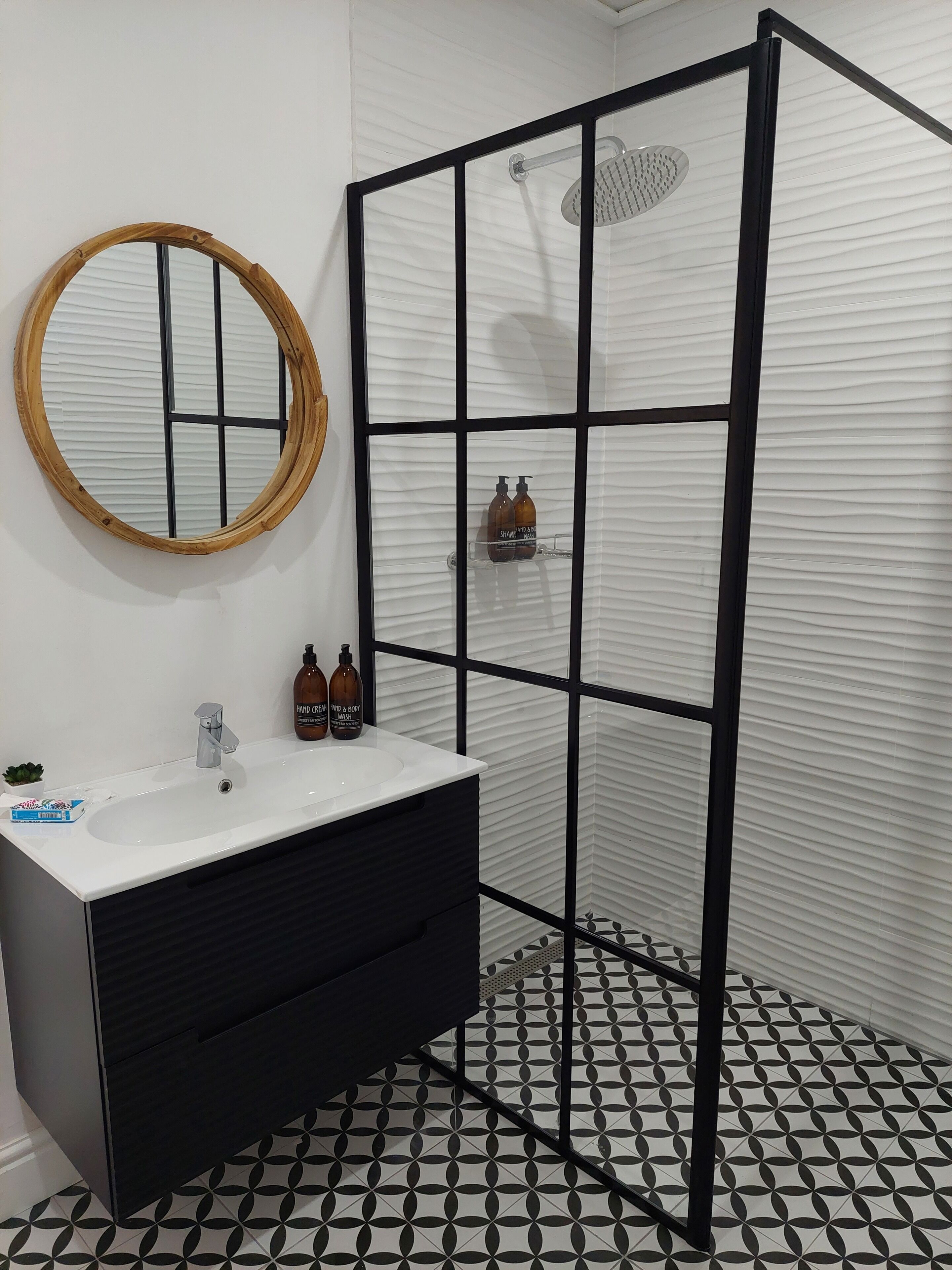 Um banheiro com piso quadriculado em preto e branco, uma pia branca com armário preto, um espelho redondo com moldura de madeira e box de chuveiro preto.