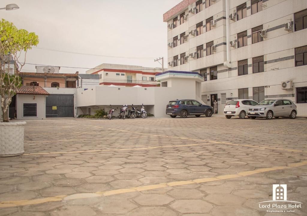 Um estacionamento com vários carros e motocicletas, um edifício com vários andares e varandas, e uma árvore em primeiro plano.