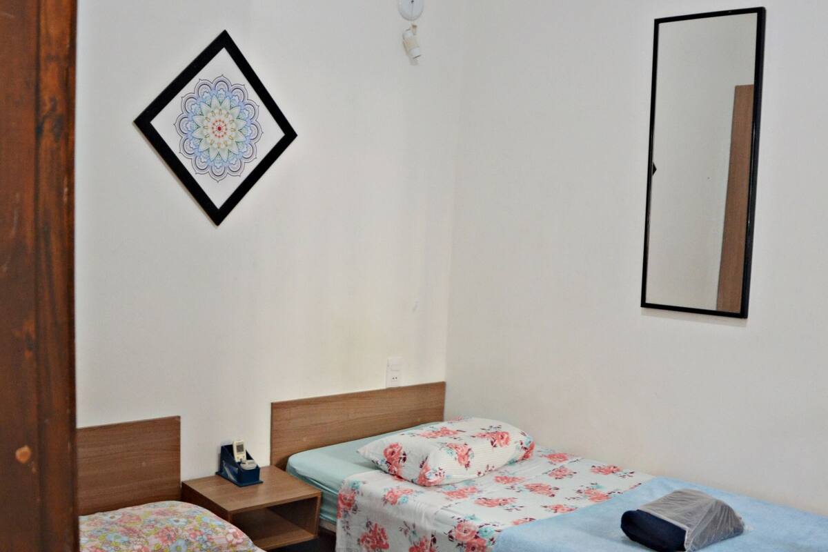 Quarto com duas camas de solteiro, cabeceira de madeira, mesa de cabeceira, quadro emoldurado, espelho e piso de madeira.