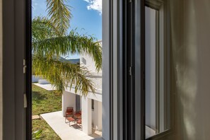Vista pela janela de um edifício moderno, com área de varanda, móveis de exterior e uma palmeira.