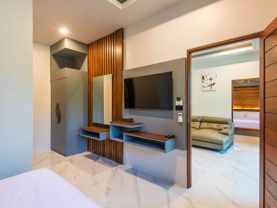 Quarto moderno com cama, TV integrada, painéis de madeira e um sofá na sala adjacente.