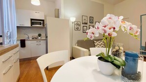 Cozinha moderna com armários brancos, micro-ondas e pia. Há uma área de refeições com uma mesa e cadeiras brancas, e um vaso de orquídeas sobre a mesa.