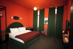 Quarto com cama, mesa de cabeceira, cadeira, escrivaninha, espelho e janela com cortinas.