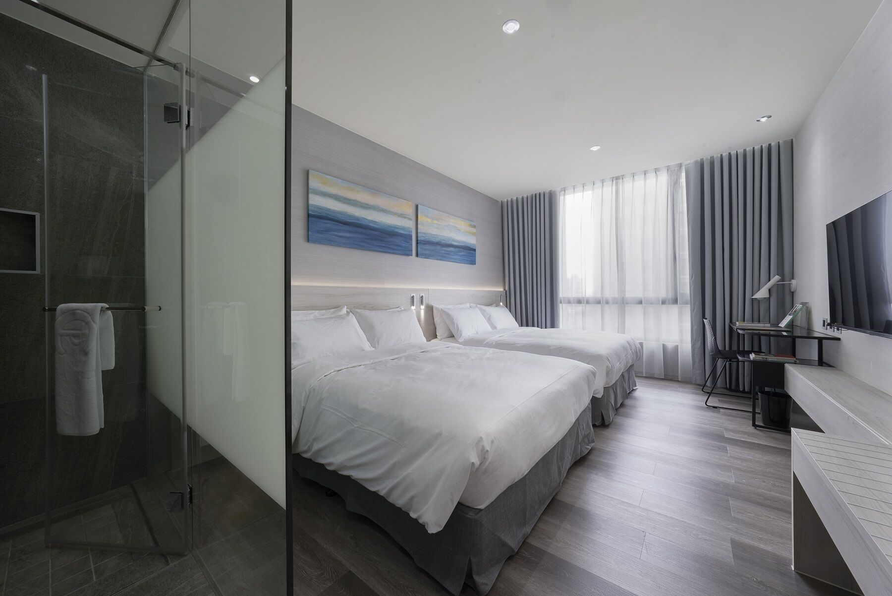 Quarto de hotel moderno com chuveiro em gabinete de vidro, duas camas, uma escrivaninha com cadeira e uma janela grande com cortinas.