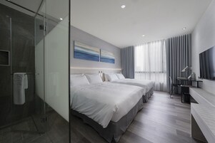 Quarto de hotel moderno com chuveiro em gabinete de vidro, duas camas, uma escrivaninha com cadeira e uma janela grande com cortinas.