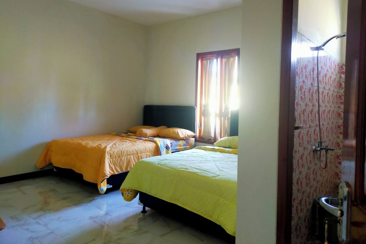 Quarto com duas camas, janela com cortinas e banheiro visível através de uma porta aberta.