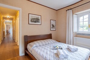 Quarto de hotel com chão de madeira, cama com colcha acolchoada, duas fotografias emolduradas na parede, janela com cortinas e corredor que dá para outro quarto.