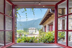 Vista através de uma janela aberta, com um edifício de pedra, vegetação e montanhas ao fundo.