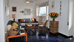 Uma sala de estar com sofá, mesa de centro e cadeira de vime, uma janela com vista e um vaso de girassóis.