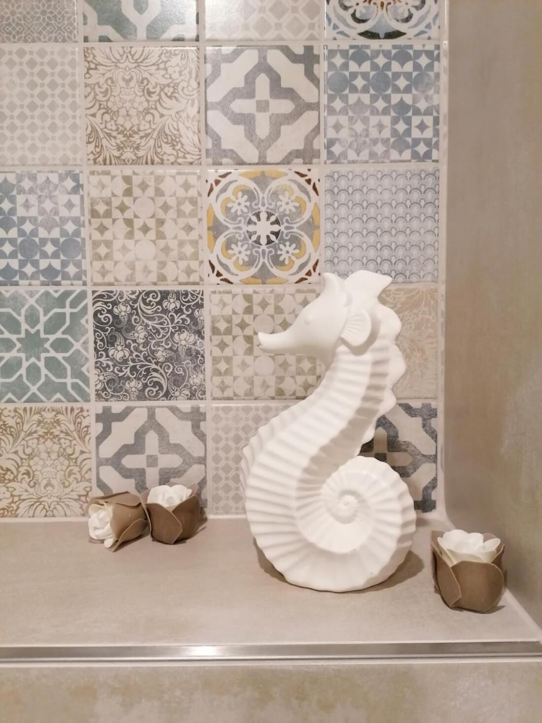 Casa de banho com parede revestida de azulejos com padrões geométricos e florais diversos. Uma escultura de cavalo-marinho branco está colocada no chão.