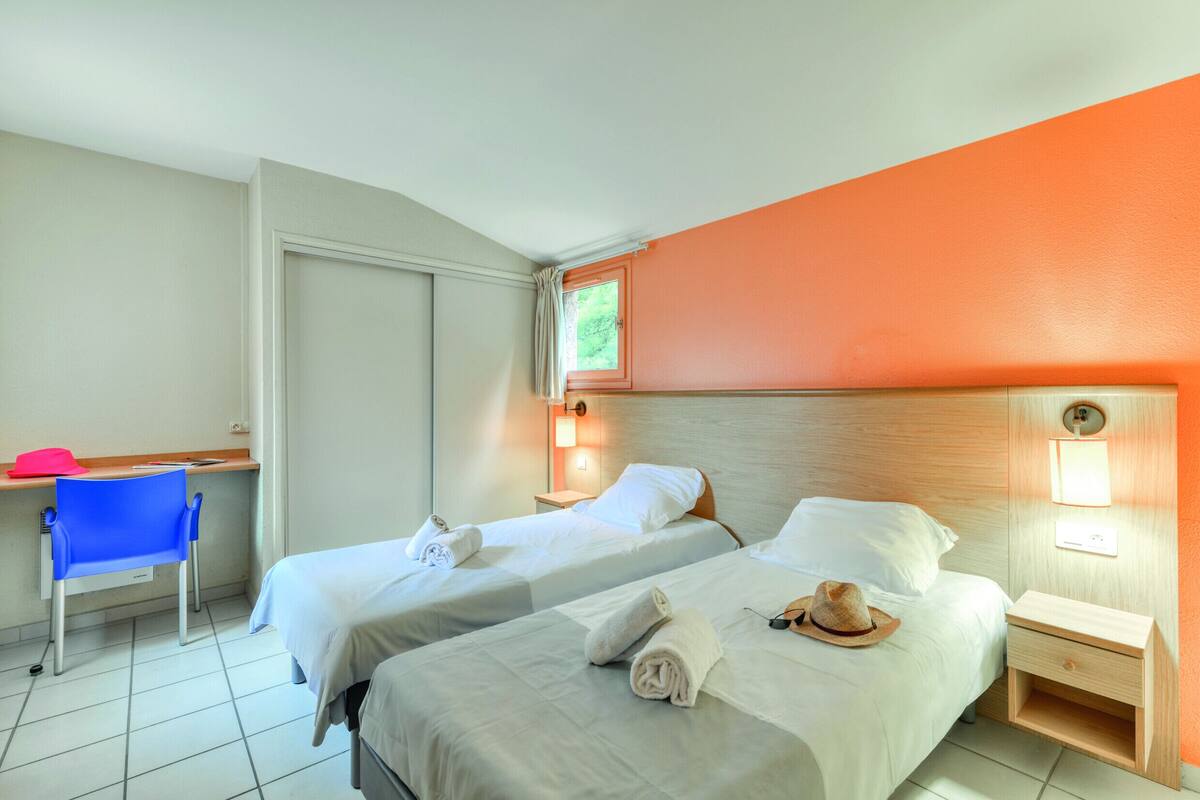 Quarto de hotel com duas camas, uma escrivaninha, uma cadeira e uma janela com cortinas.