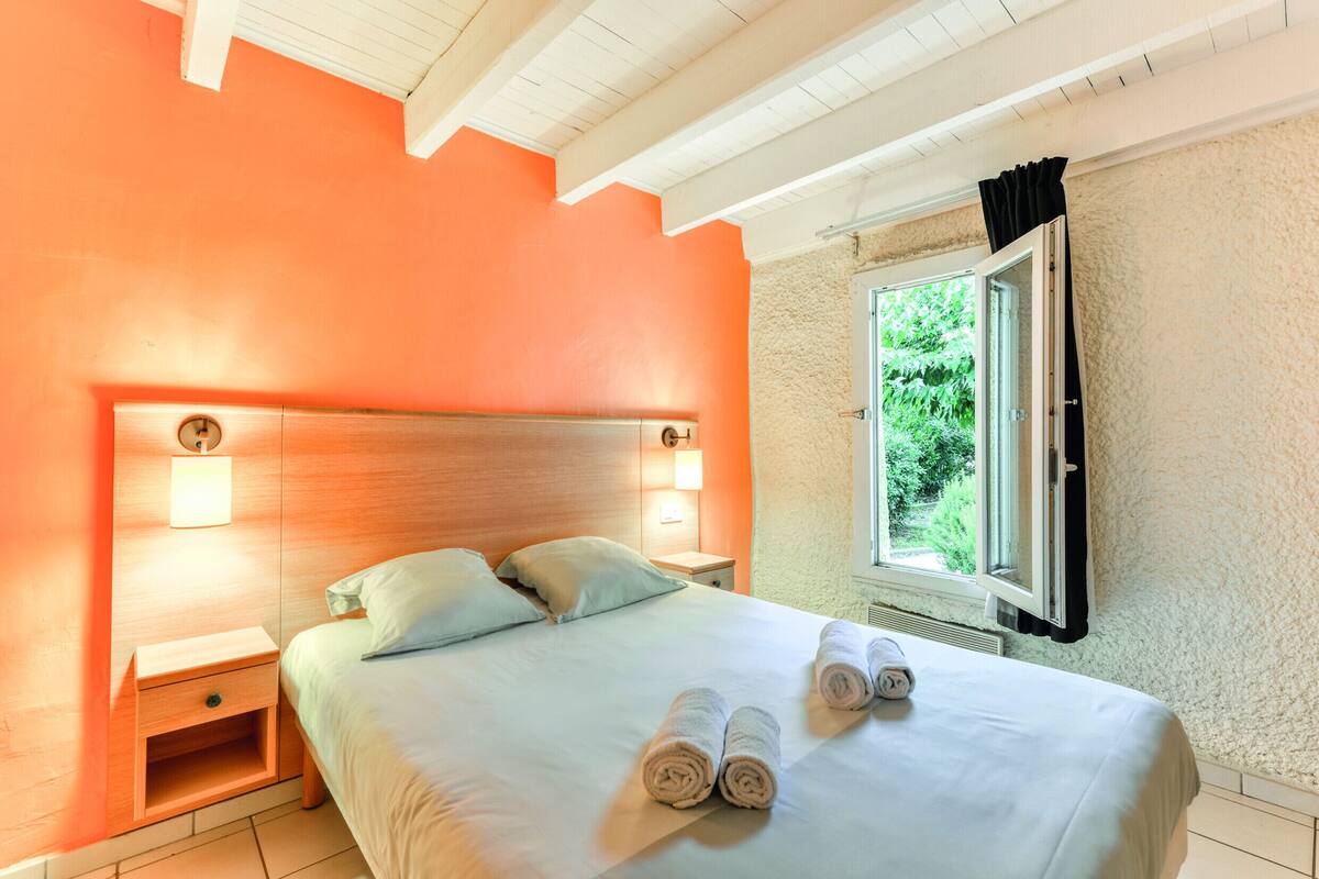 Quarto com cabeceira de madeira, duas mesas de cabeceira, janela com vista para vegetação e toalhas dobradas na cama.