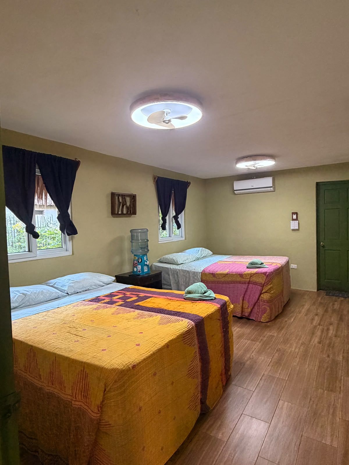 Quarto com duas camas, bebedouro, ventilador de teto e porta verde.