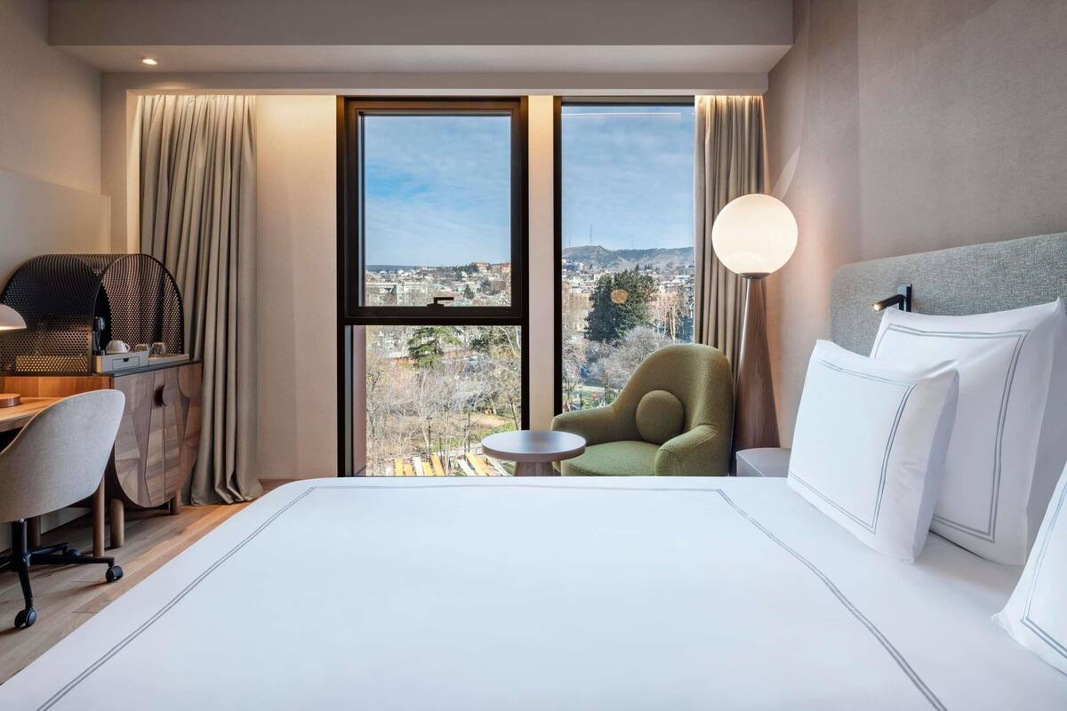 Quarto de hotel com uma cama grande, uma mesa de escritório, uma cadeira, uma mesa redonda e vista para o horizonte urbano através de grandes janelas.