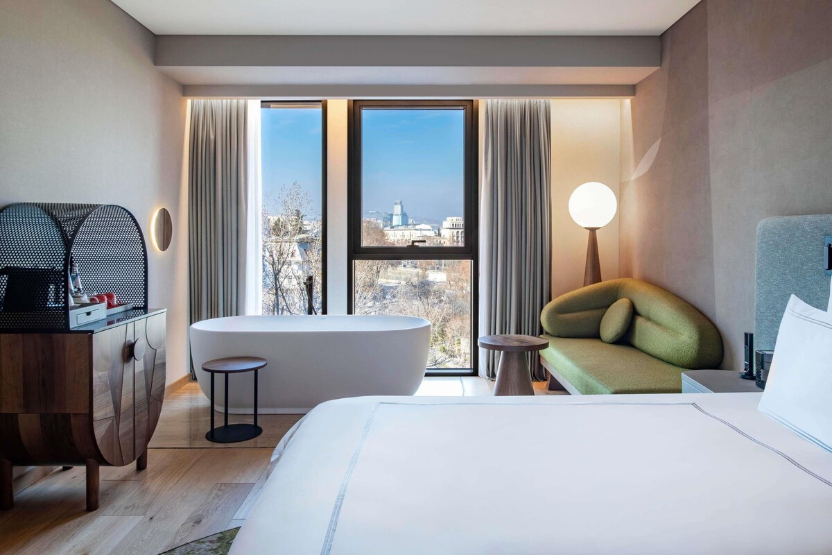 Quarto de hotel moderno com uma cama grande, uma escrivaninha, uma cadeira, uma banheira e vista para a cidade através de grandes janelas.