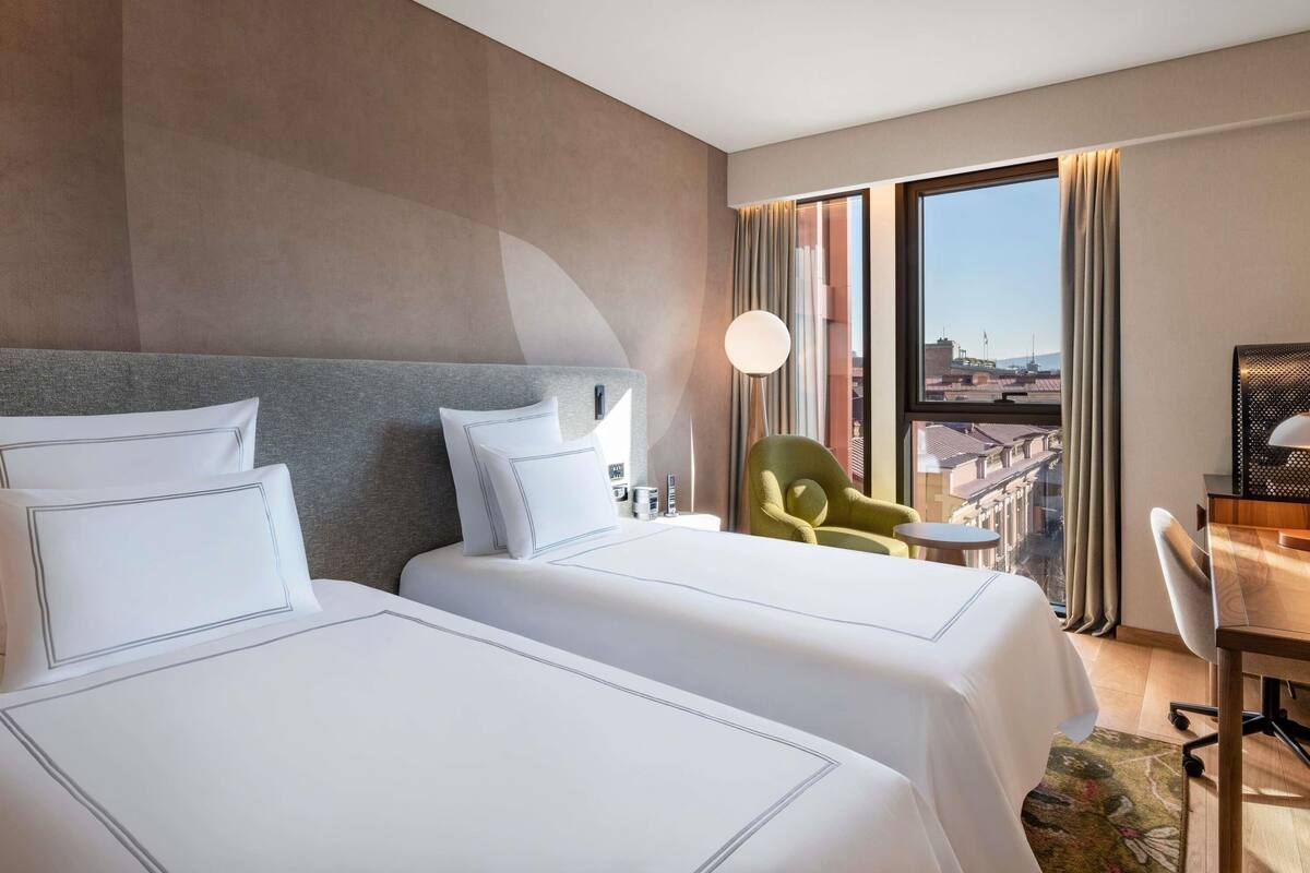 Quarto de hotel com duas camas, uma escrivaninha, uma cadeira e uma janela grande com vista para a cidade.