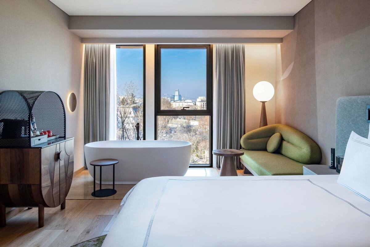 Quarto de hotel com uma cama grande, uma escrivaninha com cadeira, uma mesa redonda, uma banheira e vista para a paisagem urbana através de grandes janelas.