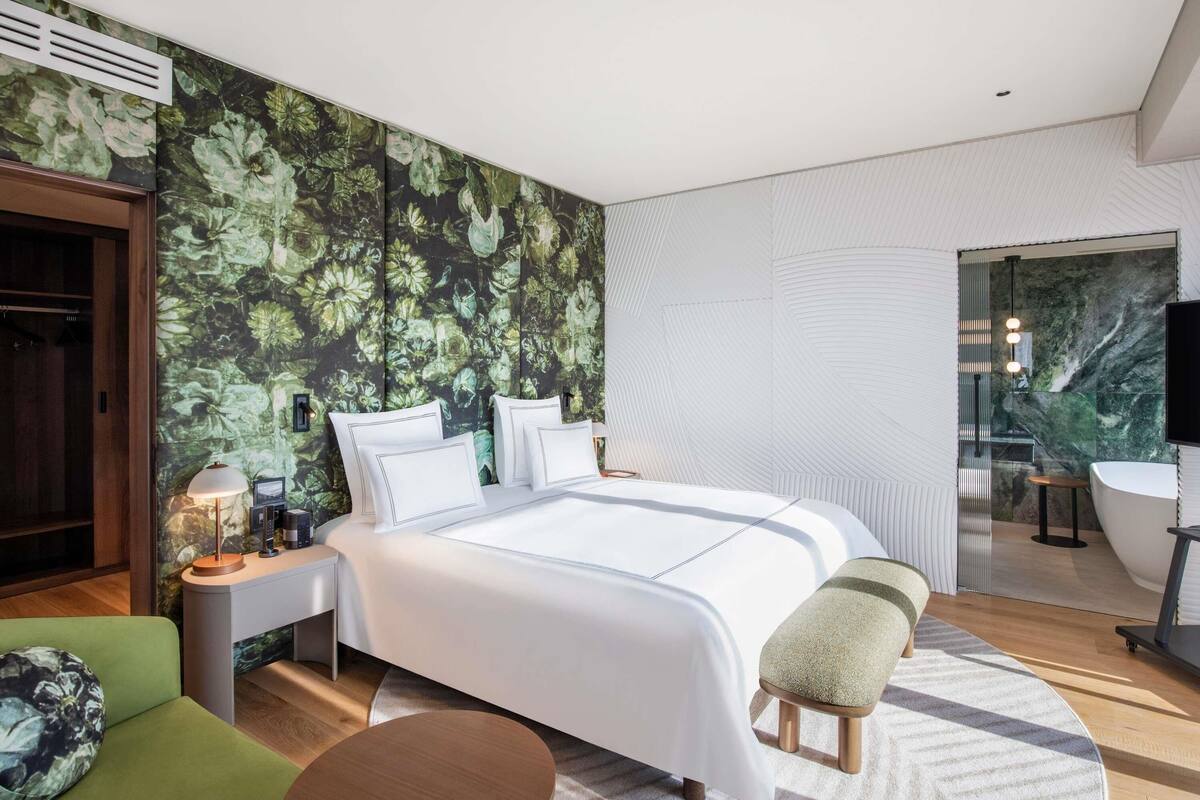 Quarto moderno com uma cama grande, uma escrivaninha, uma cadeira verde e um banheiro visível por uma porta aberta.