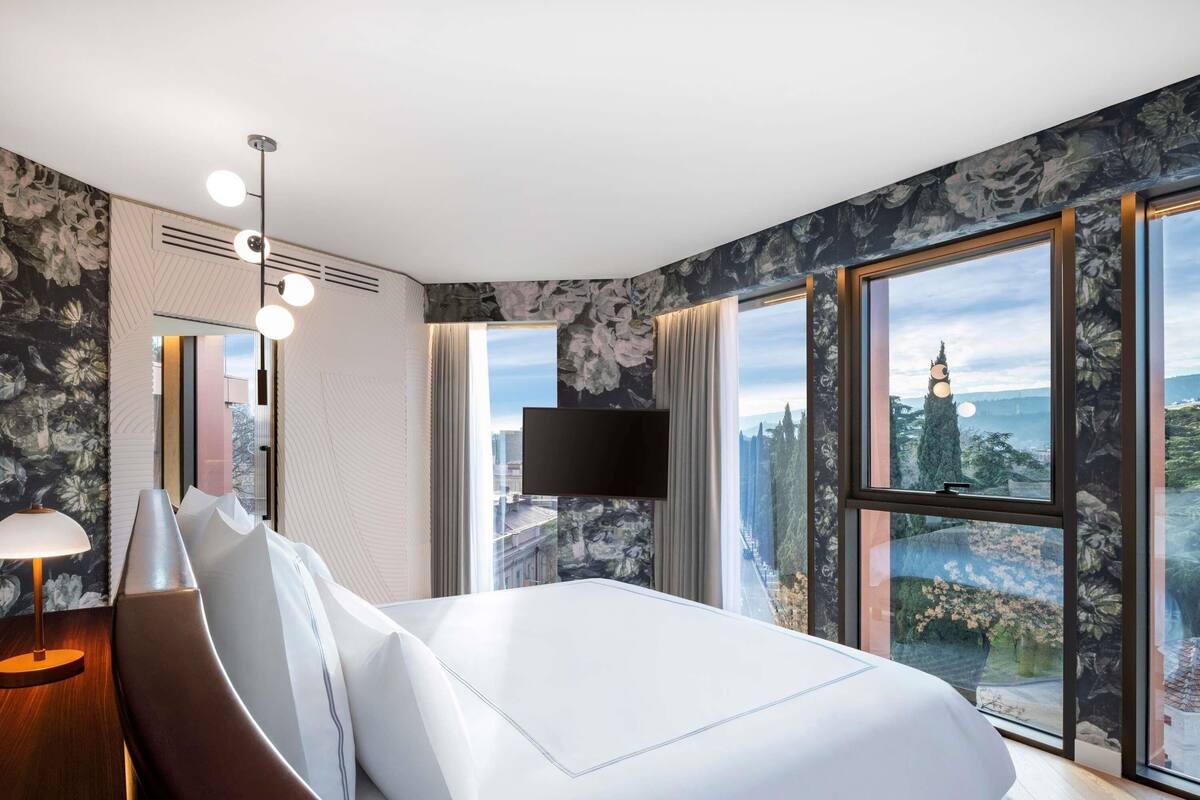 Quarto de hotel moderno com uma cama grande, televisão de tela plana, escrivaninha com abajur e amplas janelas com vista para a cidade.